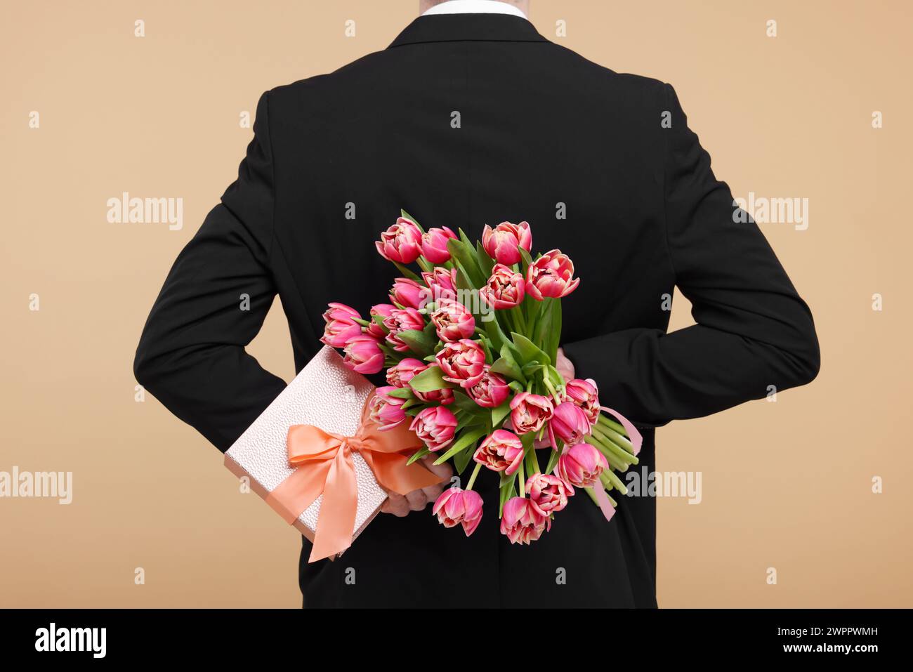 Man hiding beautiful bouquet and present on beige background, back view ...