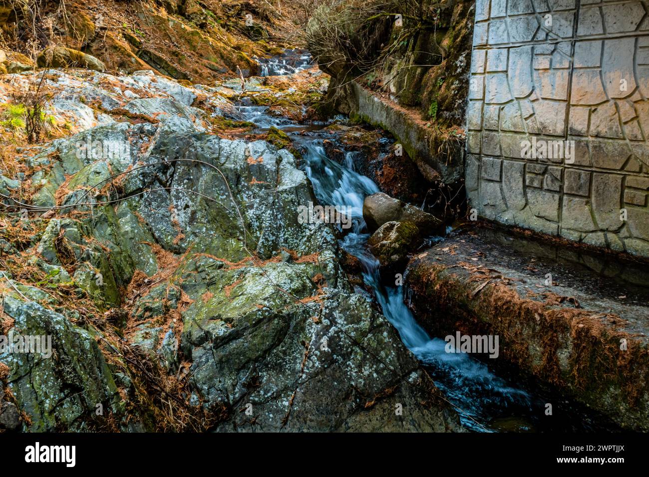 Water cascades over rocks in a stream next to a brick wall in a natural ...