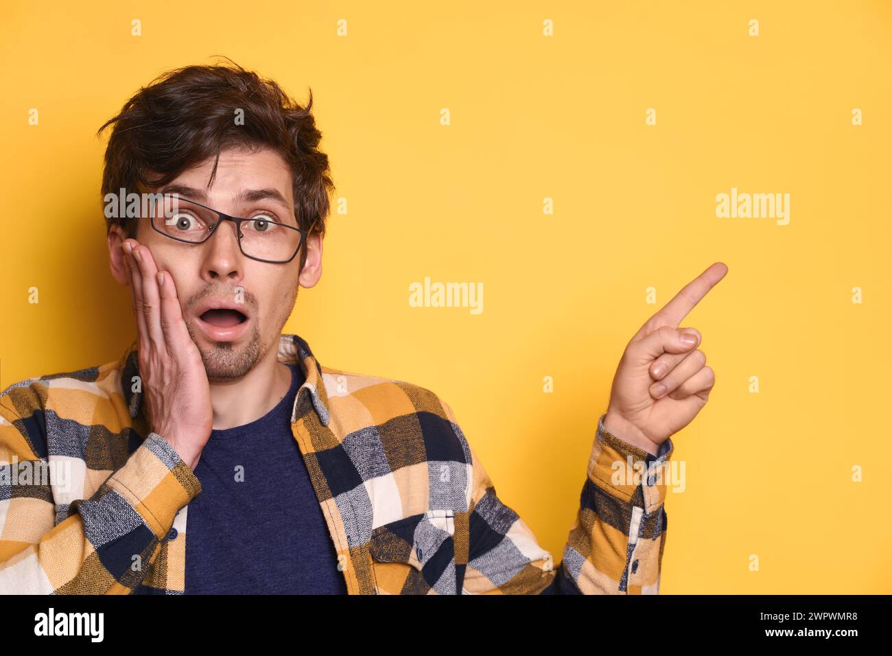 Studio portrait of shocked man in glasses and casual shirt keeps hand ...