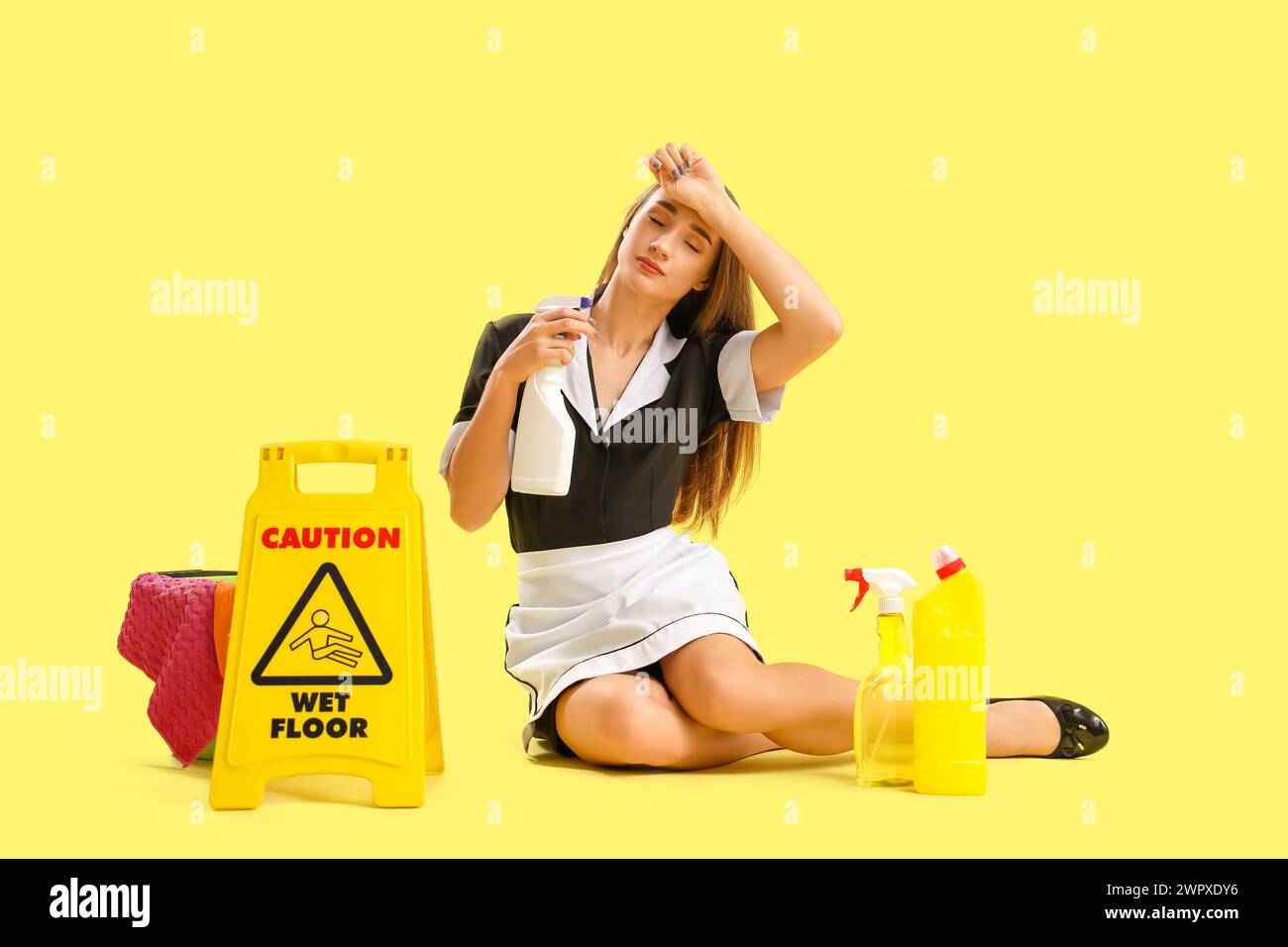 Portrait of tired female janitor with bottles of detergent and caution ...