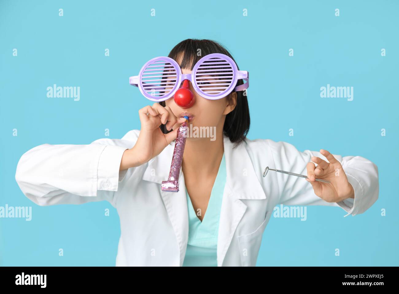Funny young female dentist with party blower and dental tool on blue ...