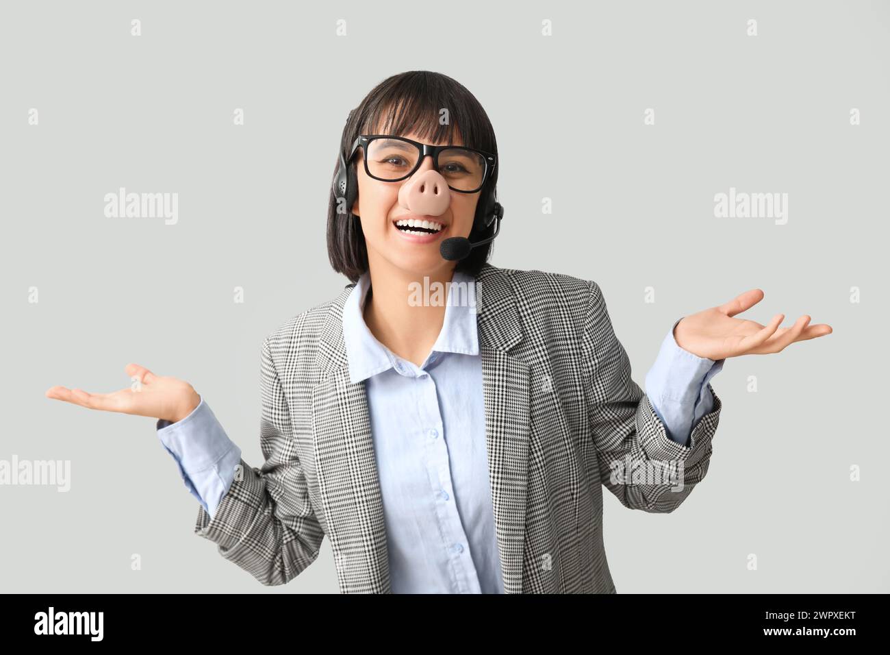 Funny young female technical support worker with headset and pig nose on grey background. Fool's ...