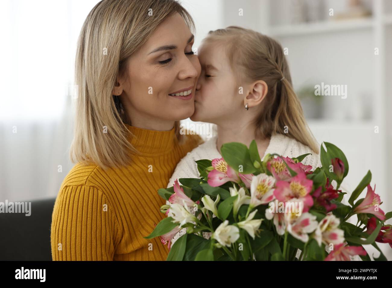 Little daughter kissing and congratulating her mom with bouquet of alstroemeria flowers at home ...