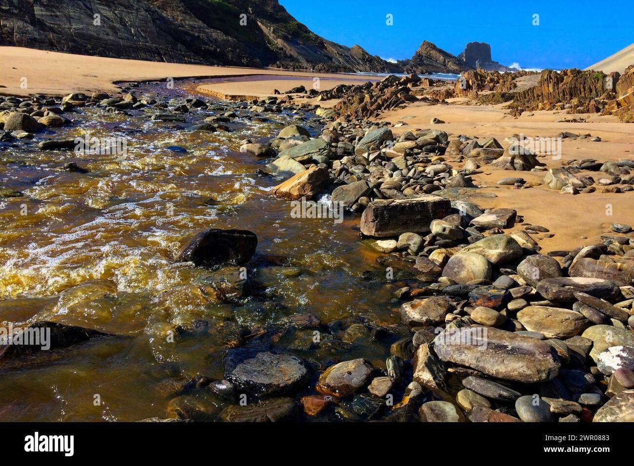 Stream with rocks leads to an open beach, flanked by towering cliffs ...