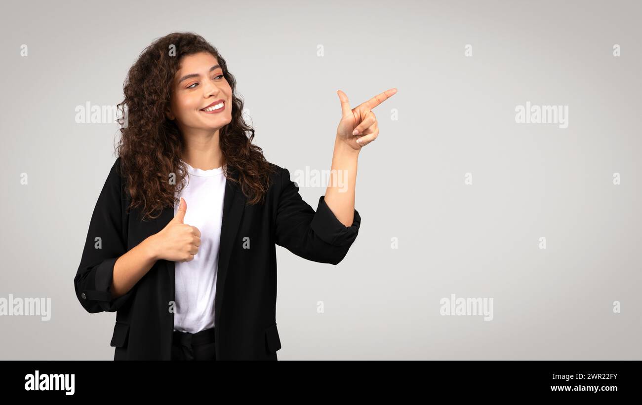 Cheerful businesswoman pointing at copy space and showing thumbs up ...