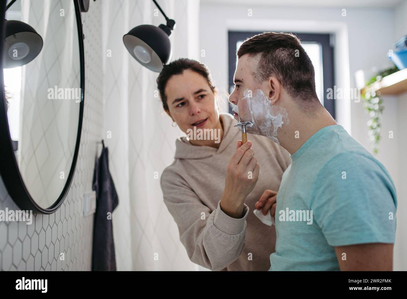 Mother applying shaving foam on son's face. Young man with down syndrome learning how to shave ...