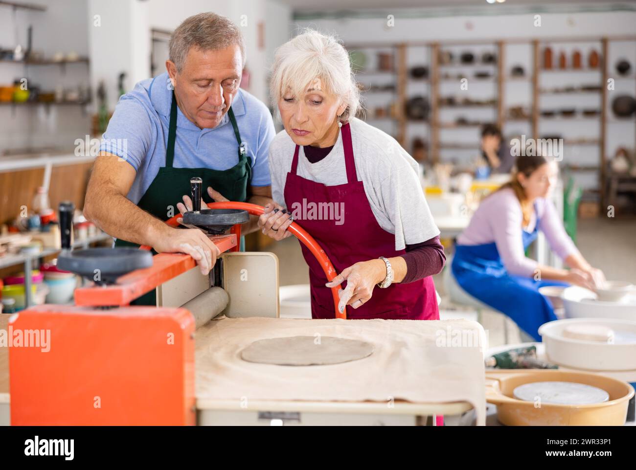 Elderly potter teaches woman how to roll clay on a craft machine clay ...