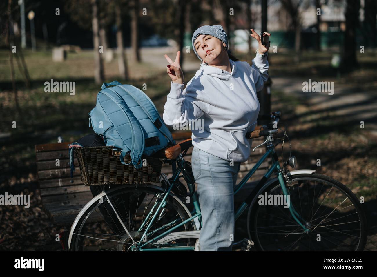 Playful young woman with bicycle making peace sign in urban park ...