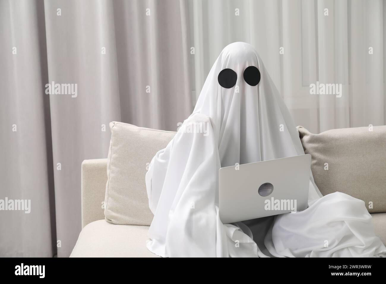 Creepy ghost. Person covered with white sheet using laptop on sofa at ...