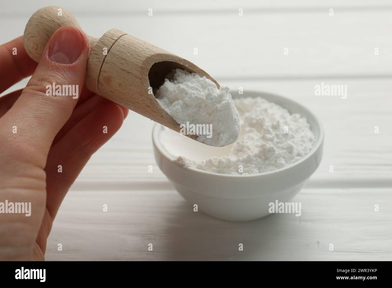 Woman taking baking powder with scoop from bowl at white wooden table ...