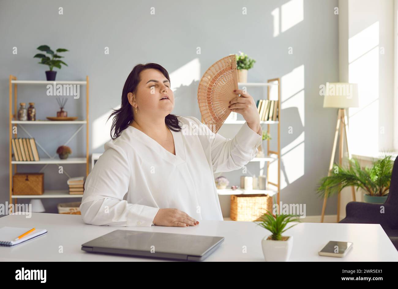 Young fat woman refreshing herself with paper fan during extreme summer ...