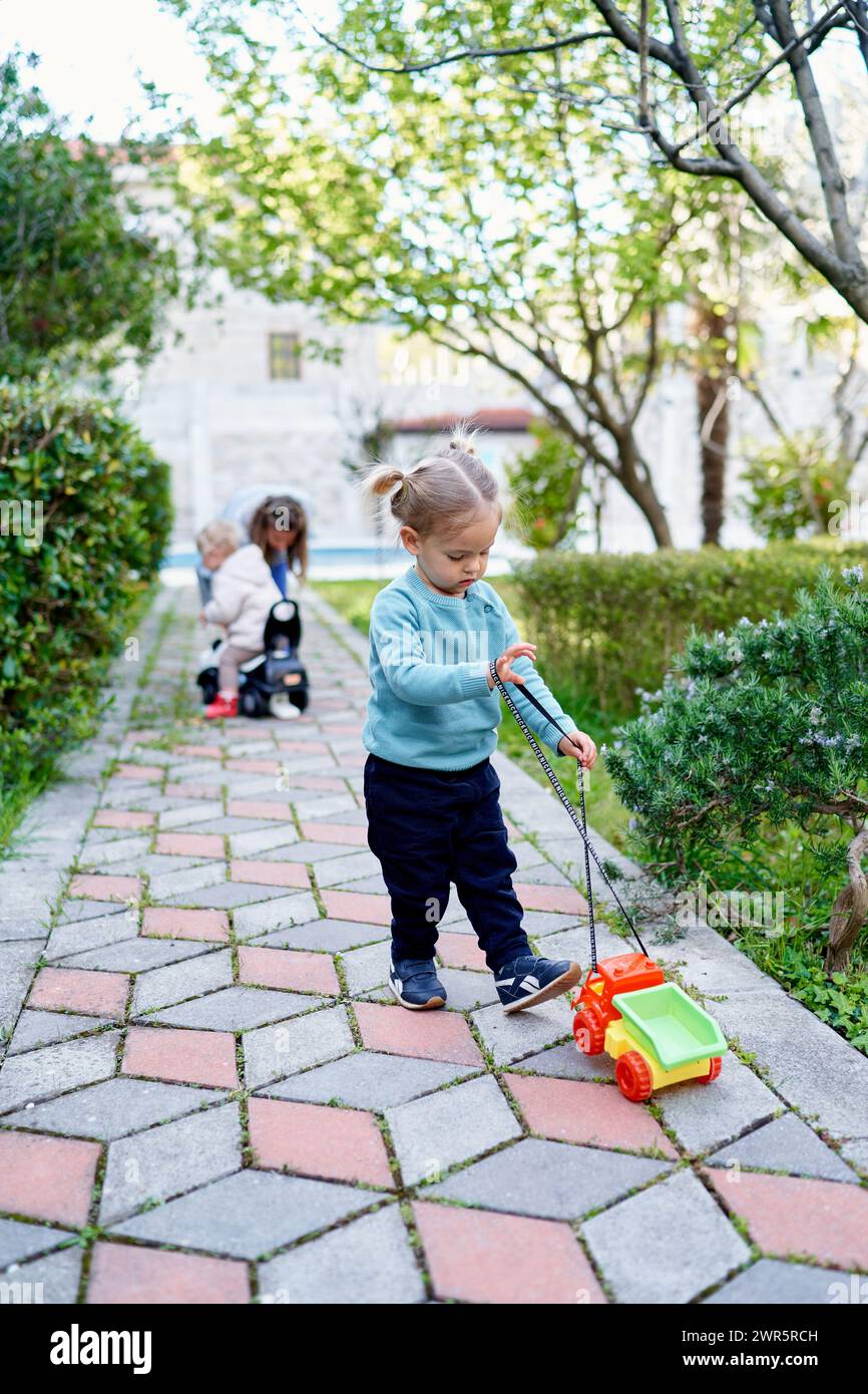 Little girl with a colorful toy car on a string stands on a paved path ...