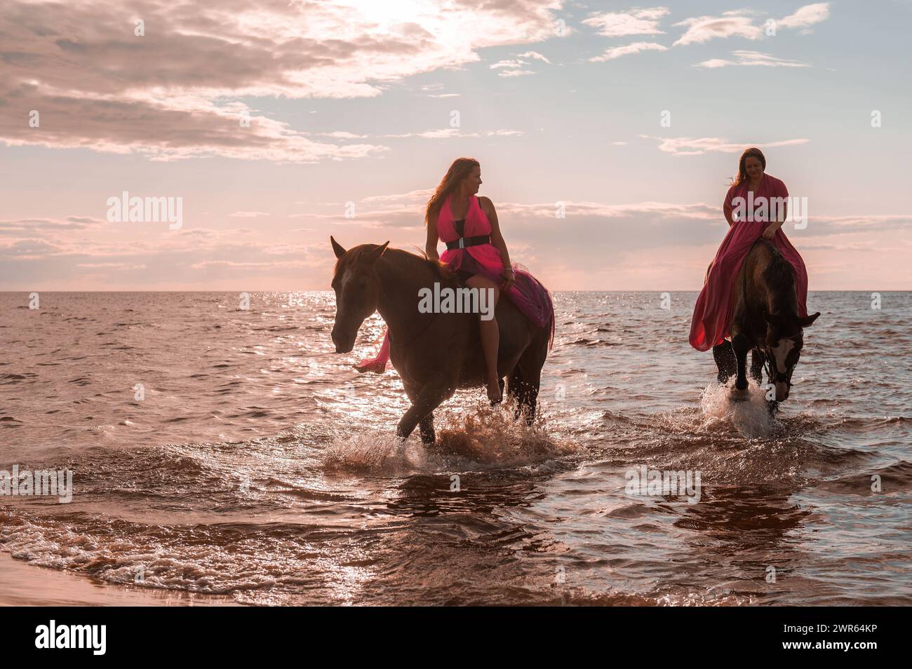 Two women horseback riding at sunset on a beach, heading into the ocean ...