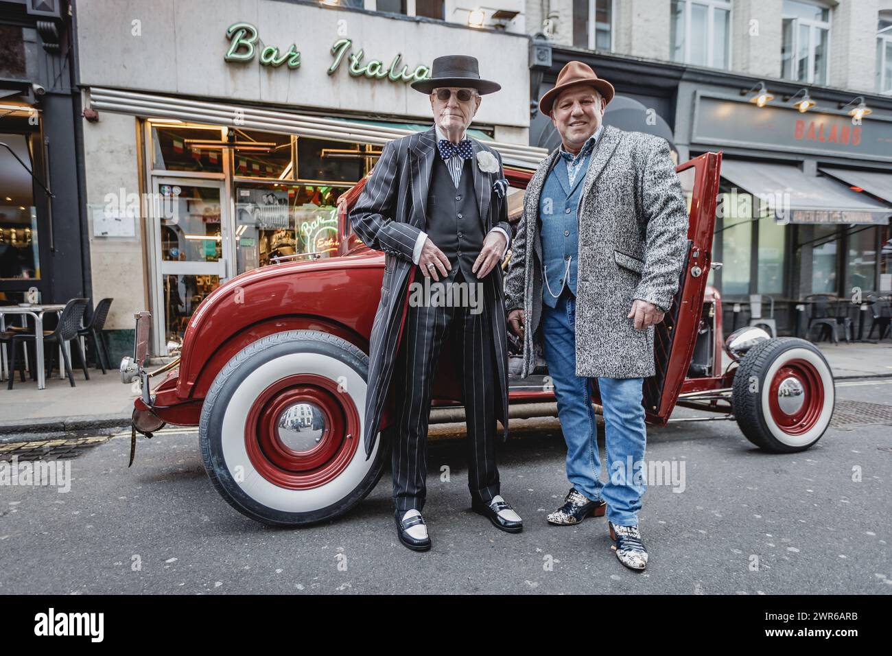 Two well-dressed gentlemen stand by a classic car outside Bar Italia in ...