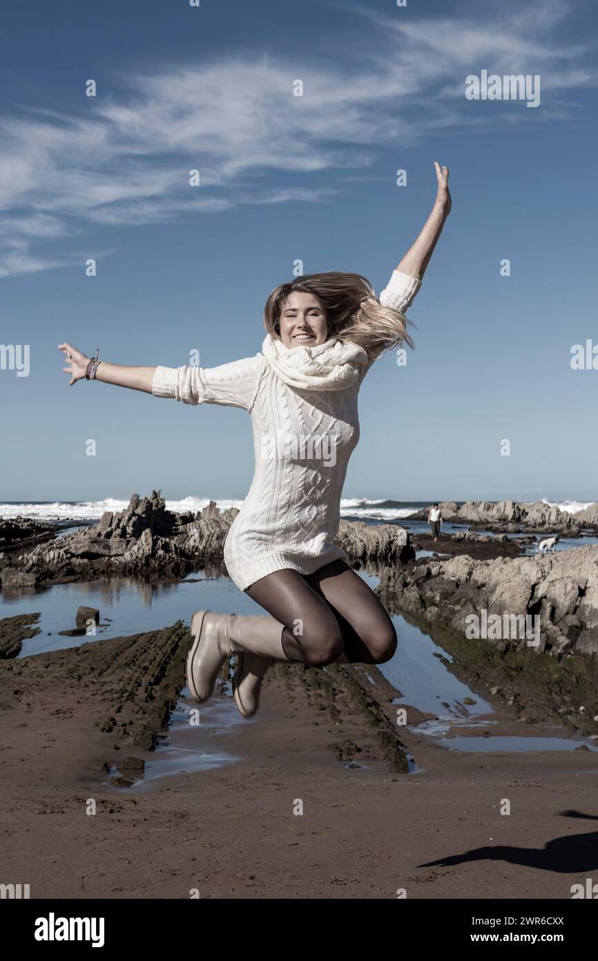 A woman is jumping in the air on a beach. She is wearing a white dress ...
