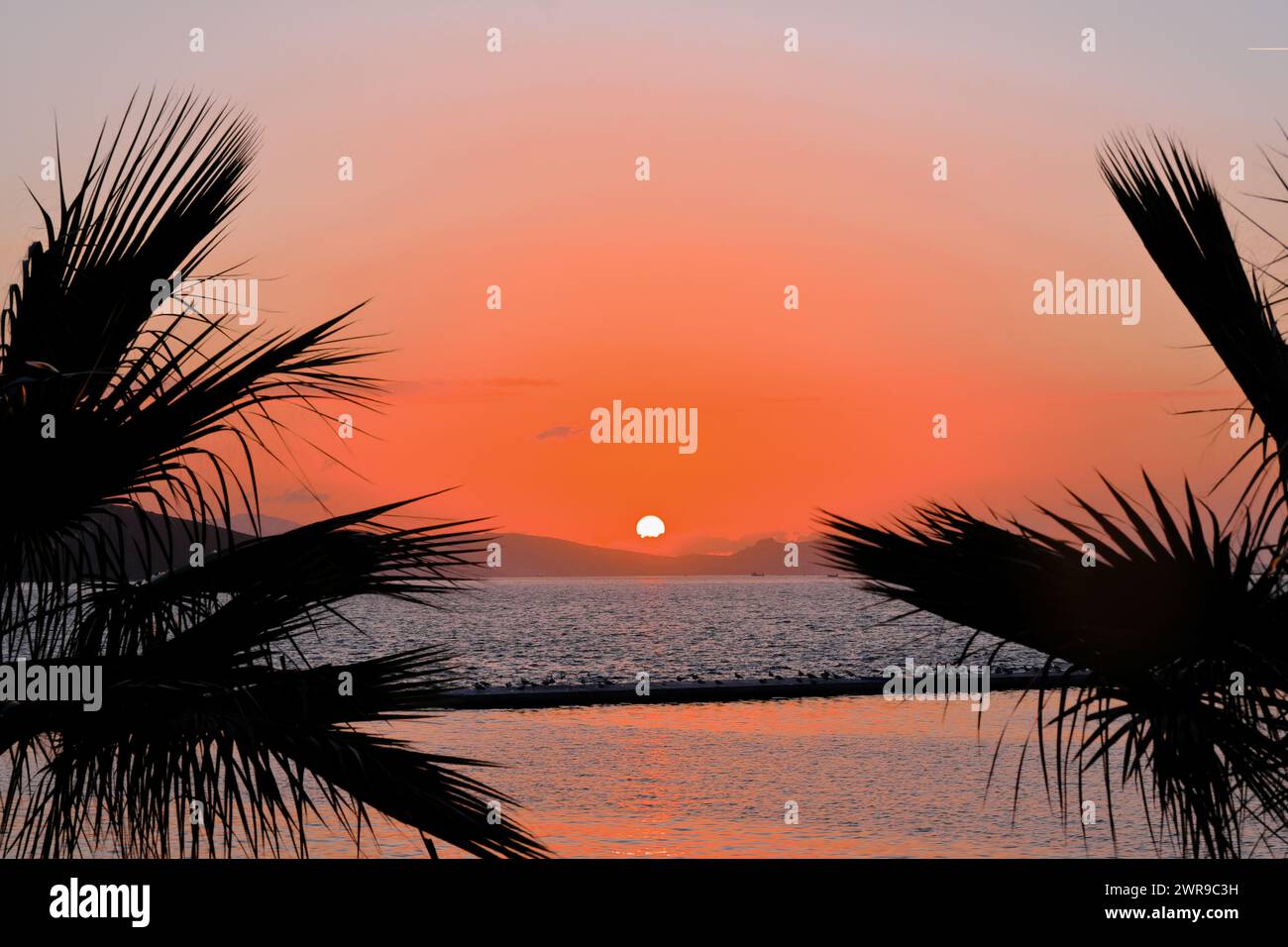 Crimson sky and full sphere sun at sunset by the sea behind the hills ...