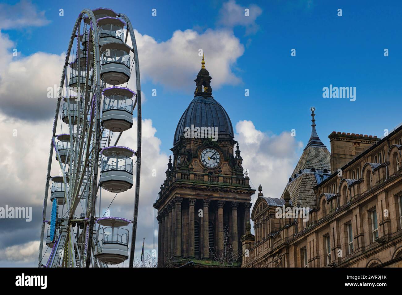 Ferris wheel beside a historic clock tower under a blue sky with clouds ...