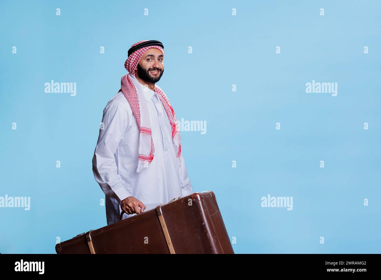 Smiling muslim traveler holding vintage leather suitcase and looking at ...