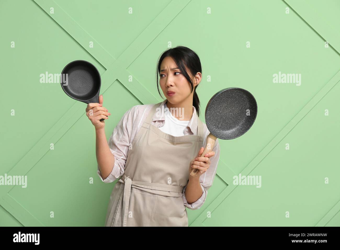 Portrait of doubtful Asian woman with frying pans on green background ...