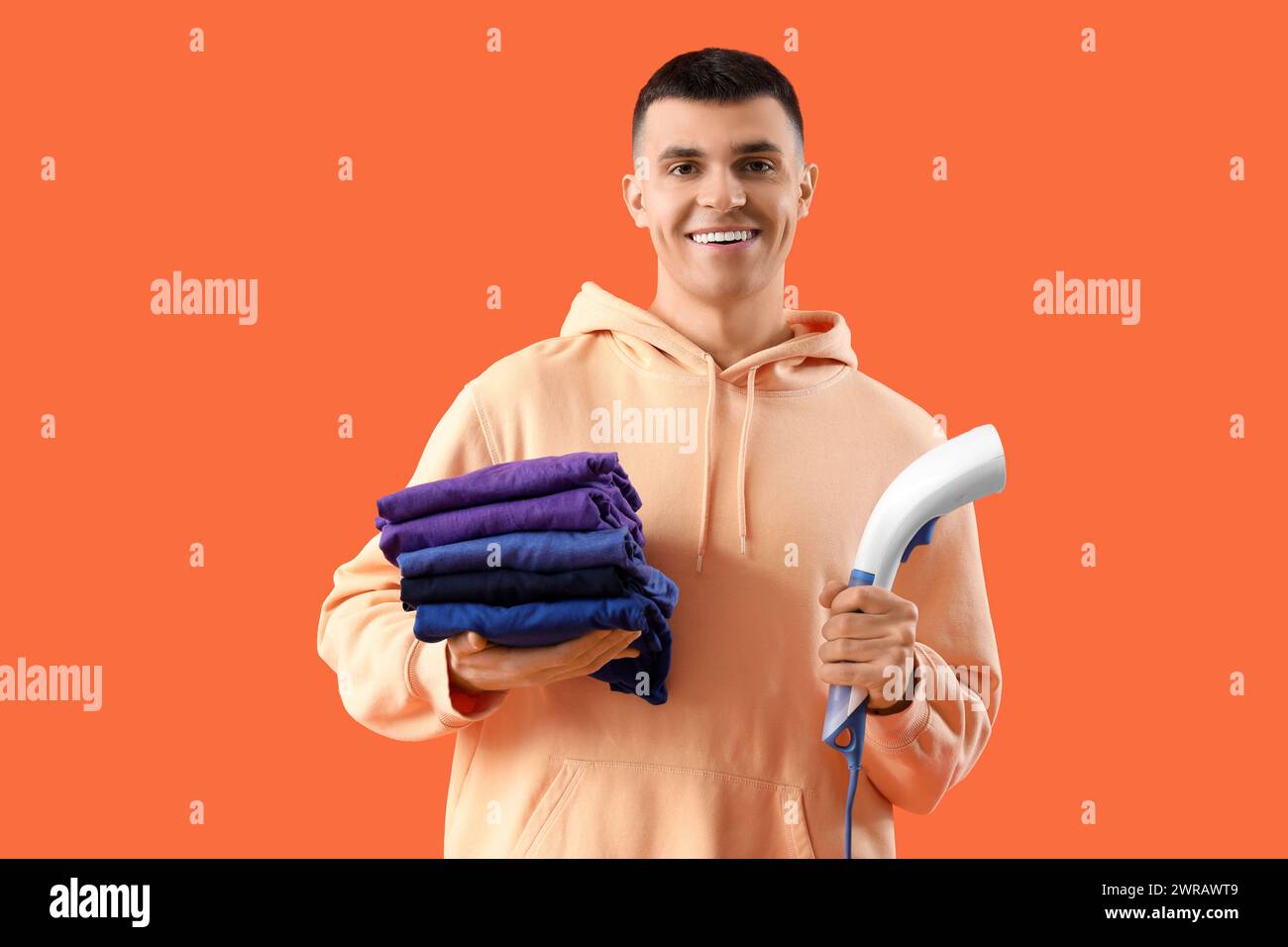 Happy young man with steamer and stack of clean clothes on orange ...