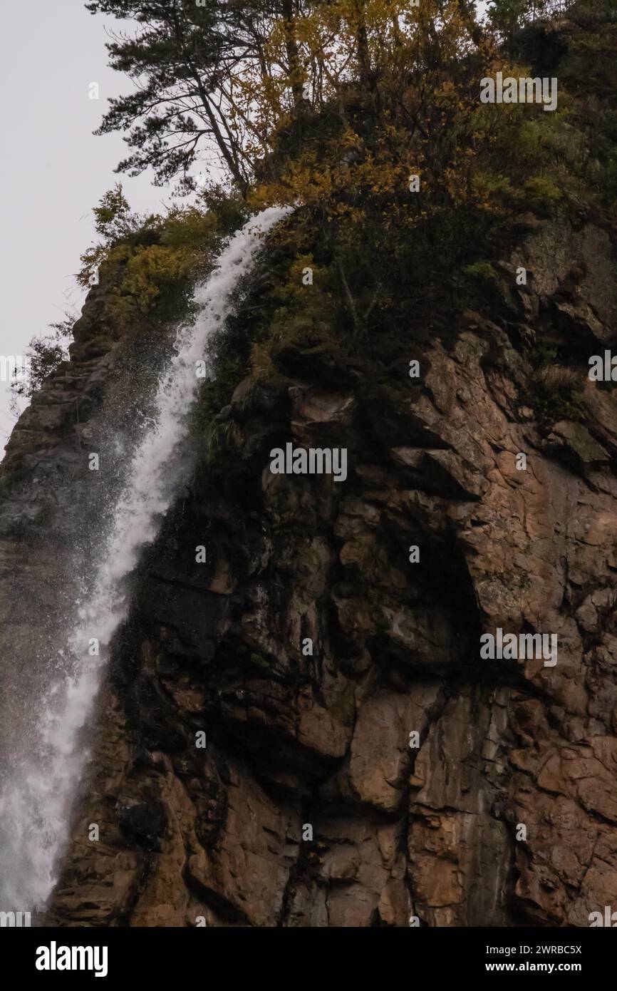 Vertical cliff face with a slender waterfall and scattered autumnal ...