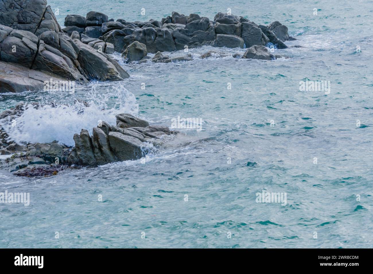 Waves crash onto boulders on the shore under a blanket of clouds, in ...