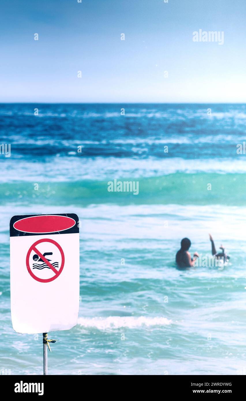 A white sign with a no swimming symbol stands at a beach, warning ...