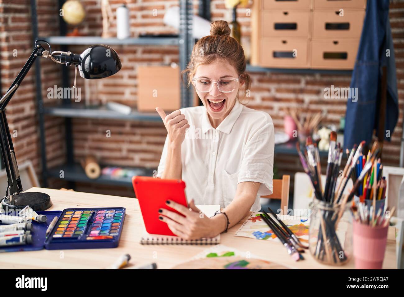 Cheerful young blonde artist woman joyfully pointing thumb to the side ...