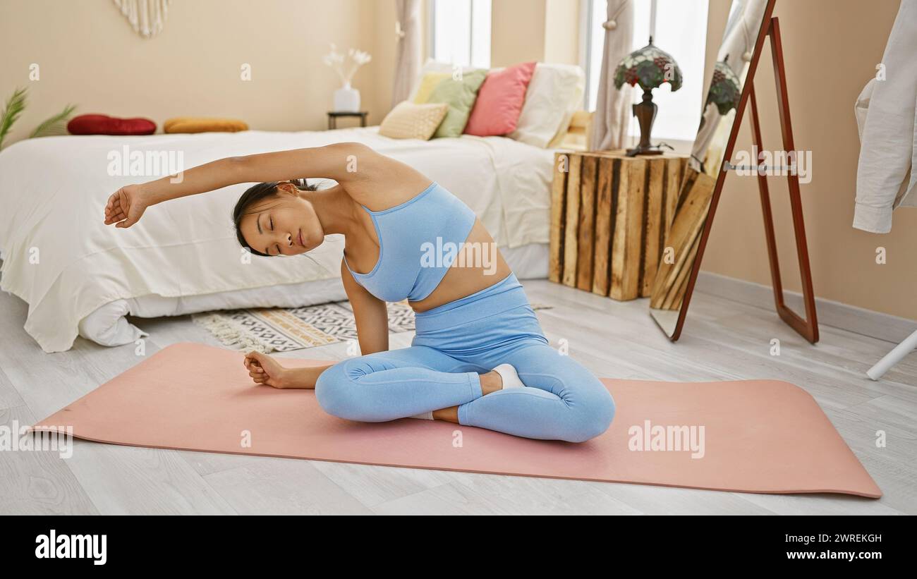 A young asian woman stretches on a yoga mat in a cozy bedroom setting ...