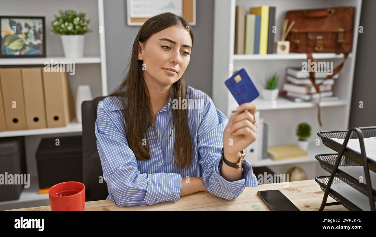 Young hispanic woman contemplating a credit card in a modern office ...