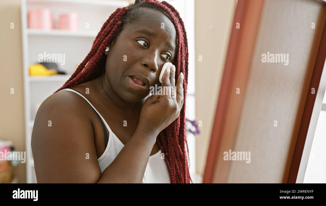 A surprised black woman with braids examines her face in a mirror in a ...