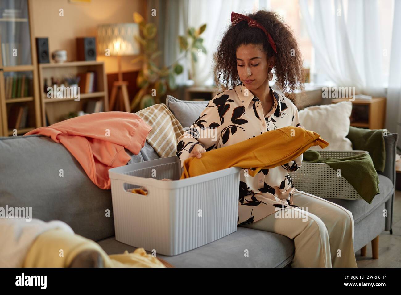 Portrait of young African American woman folding and putting away clean clothes on laundry day ...
