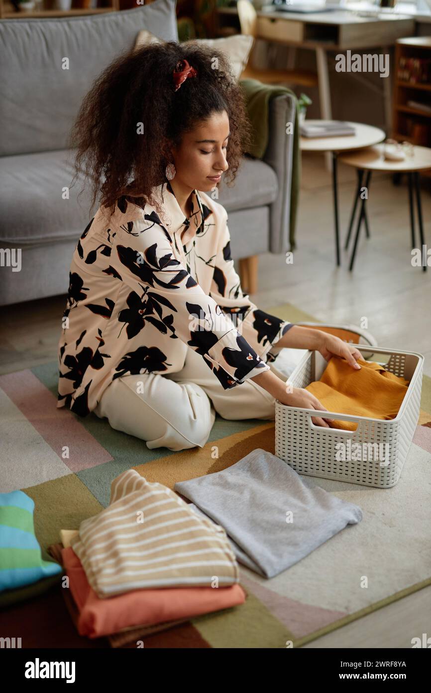 Full length portrait of young woman folding and sorting clean clothes ...