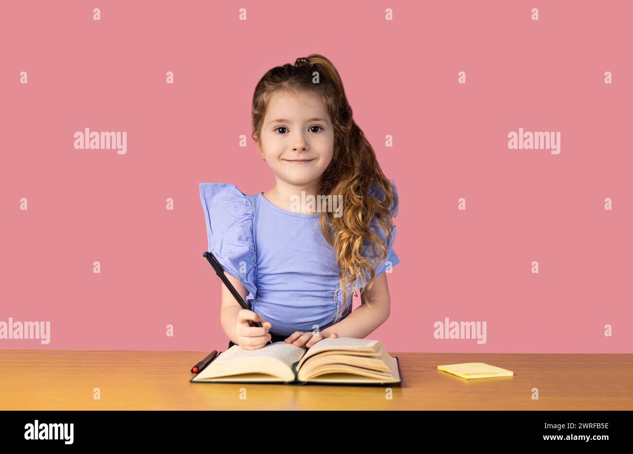 A little girl writes in a notebook and holds a pen in her right hand ...