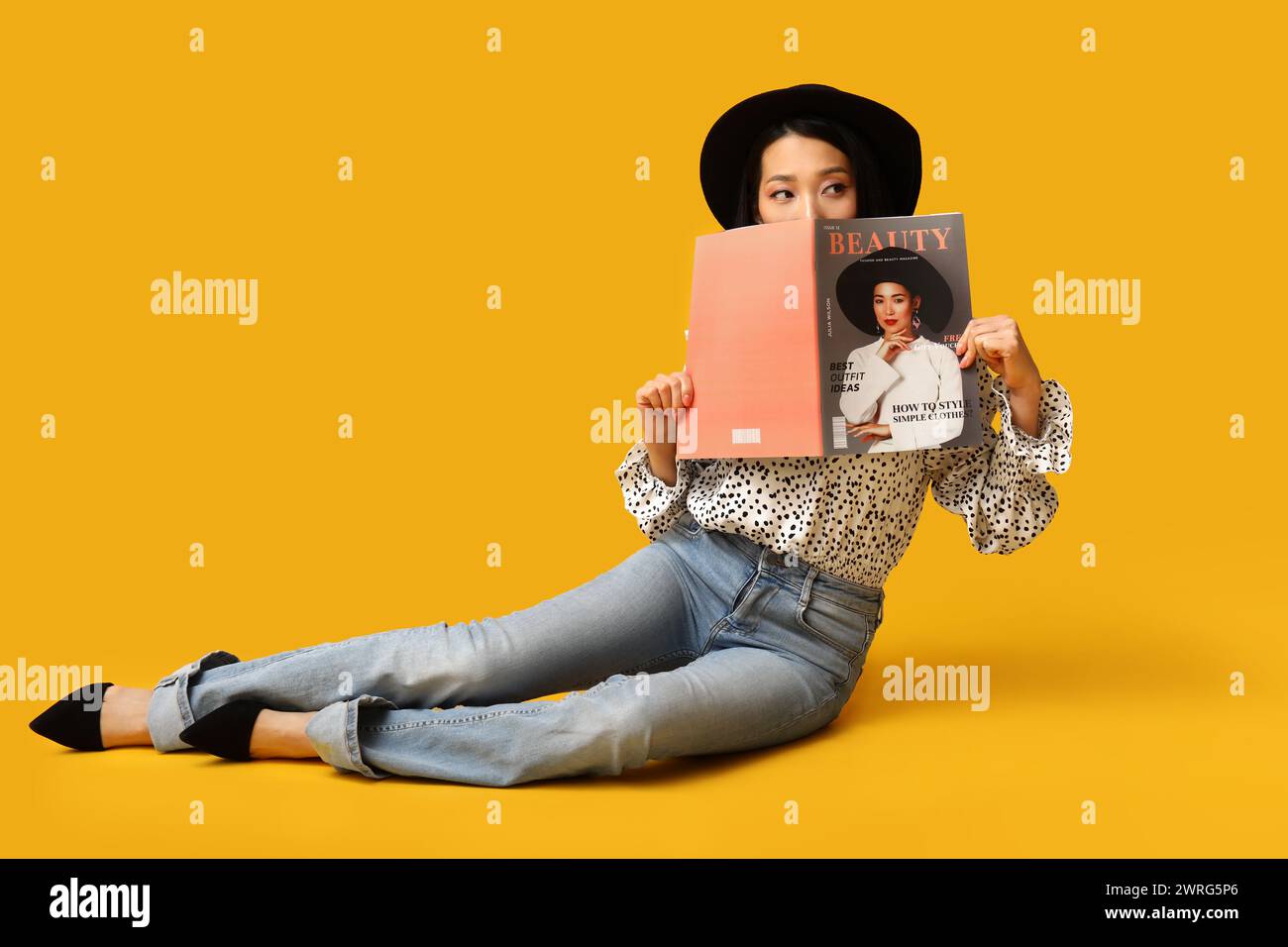 Beautiful young Asian woman in stylish jeans with magazine sitting on ...