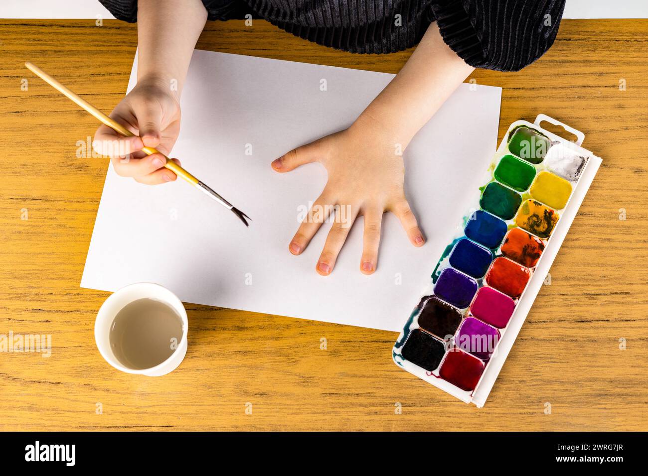 A child is using watercolors to paint a hand on a piece of paper placed ...