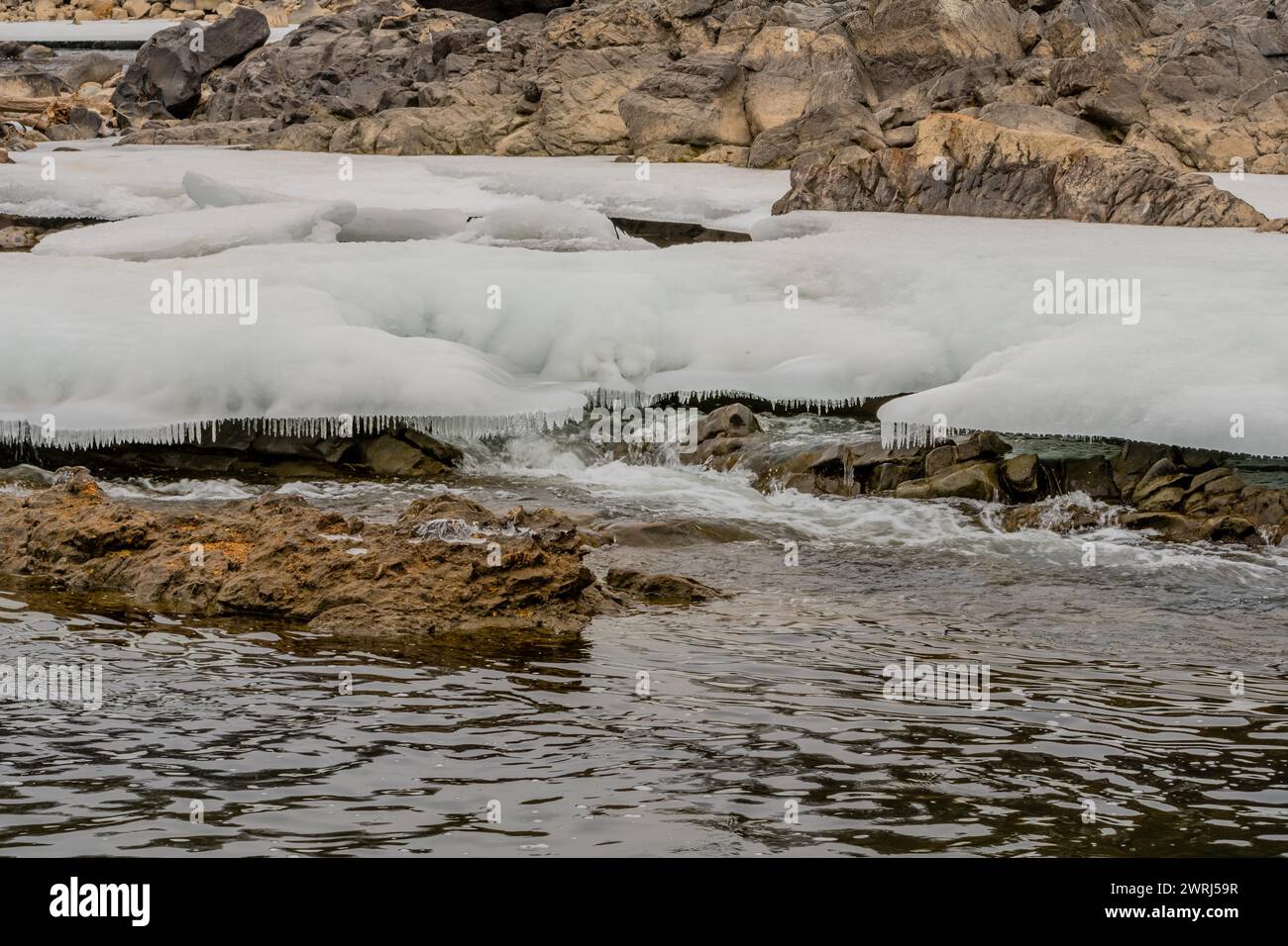 Packed snow and ice with ice-cycles that look like teeth over rapidly flowing water in river ...