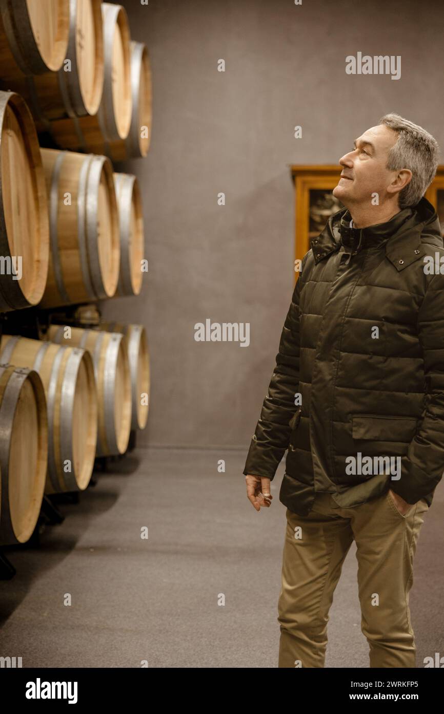 A thoughtful winemaker stands among oak barrels, appreciating the aging ...