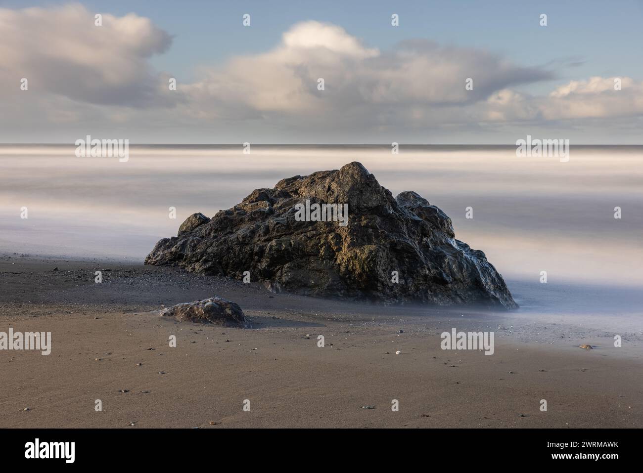 A large rock sits on a beach, with the ocean in the background. The ...