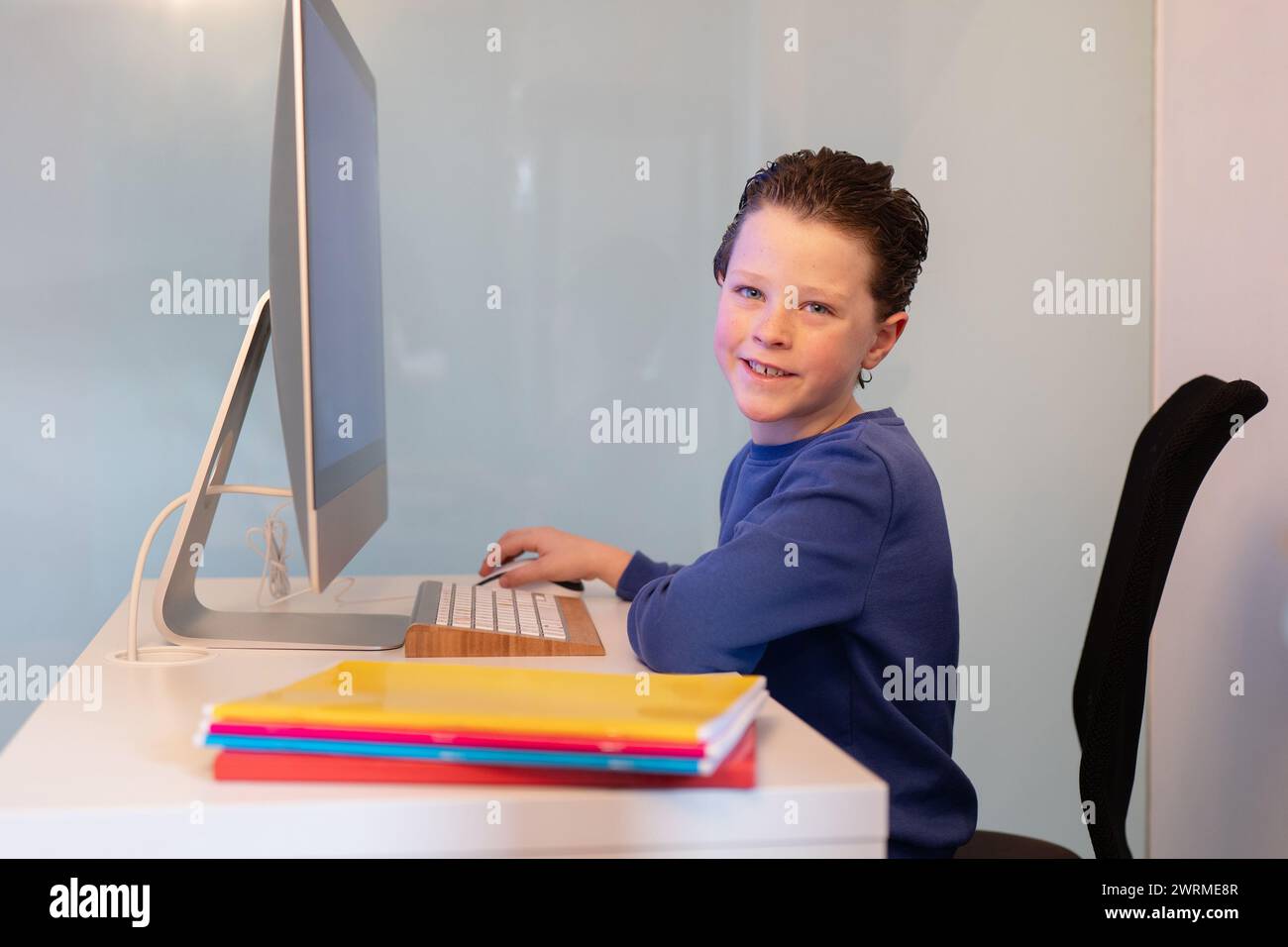 A happy child with a bright smile sits at a desk, using a computer with ...