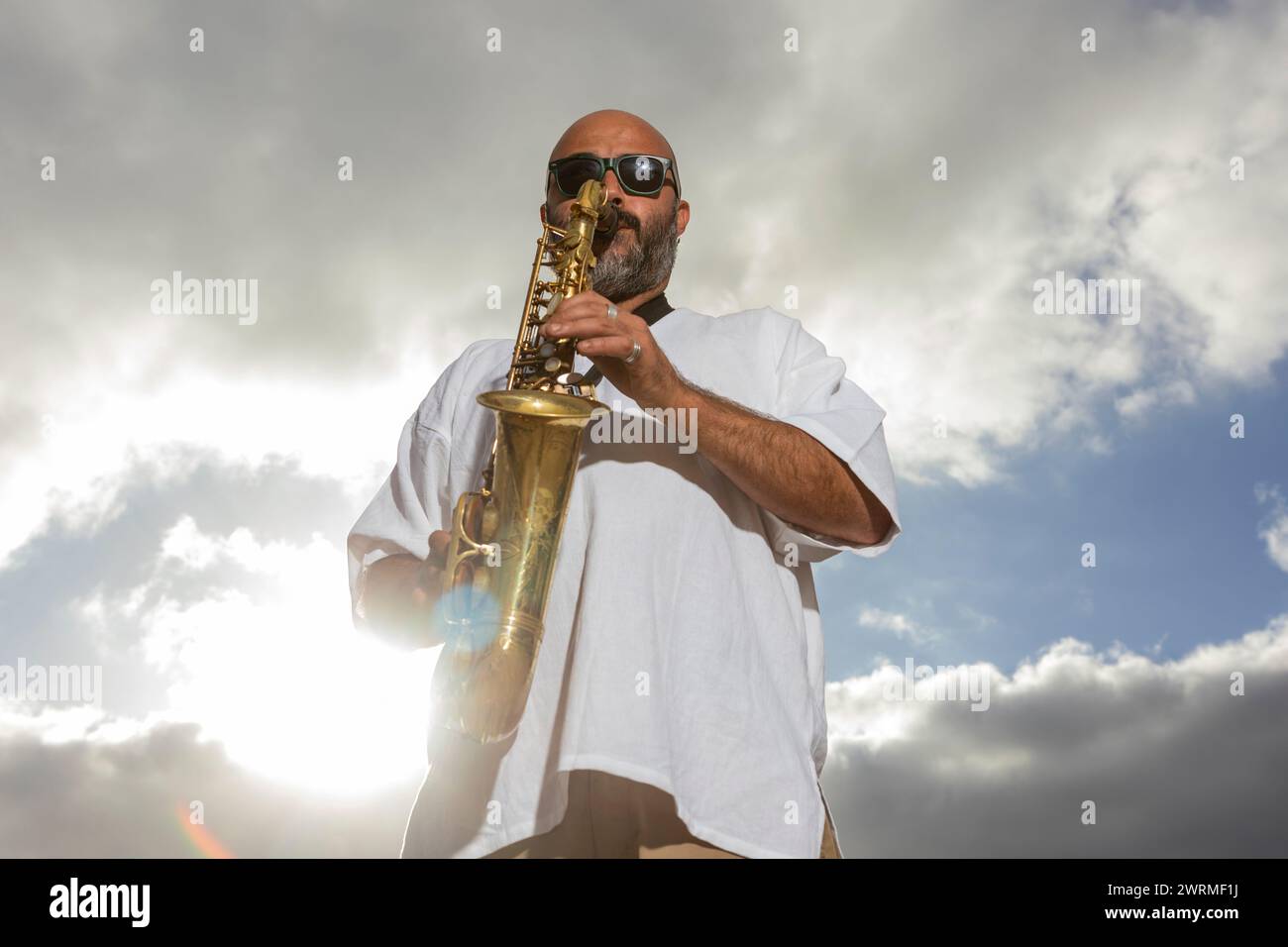 From below male saxophonist performs under a cloudy sky with sunlight ...