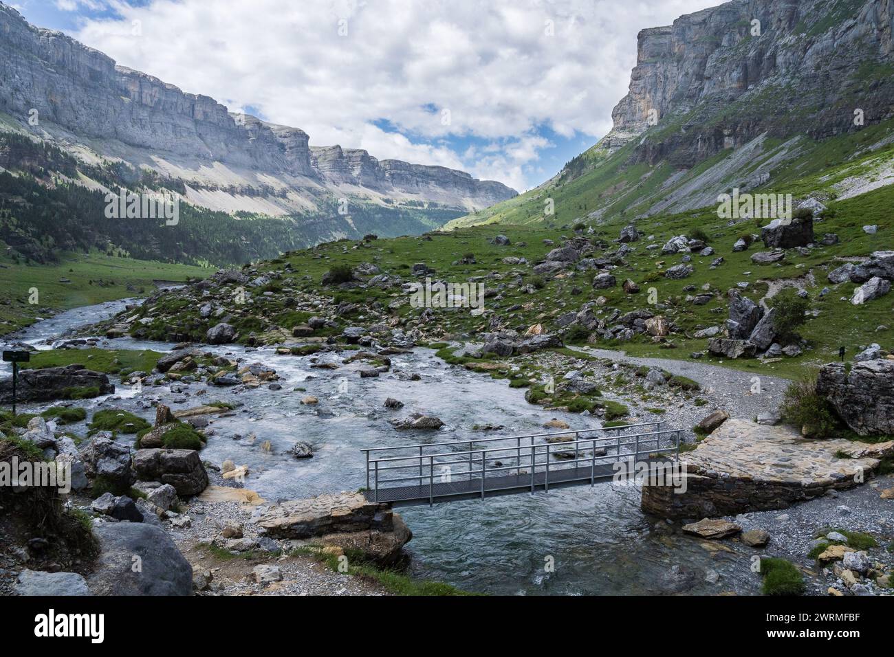 A tranquil river flows through the Ordesa Valley with a simple bridge ...
