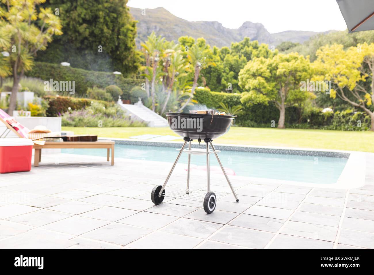 A charcoal grill is ready for a barbecue by a poolside with lush ...
