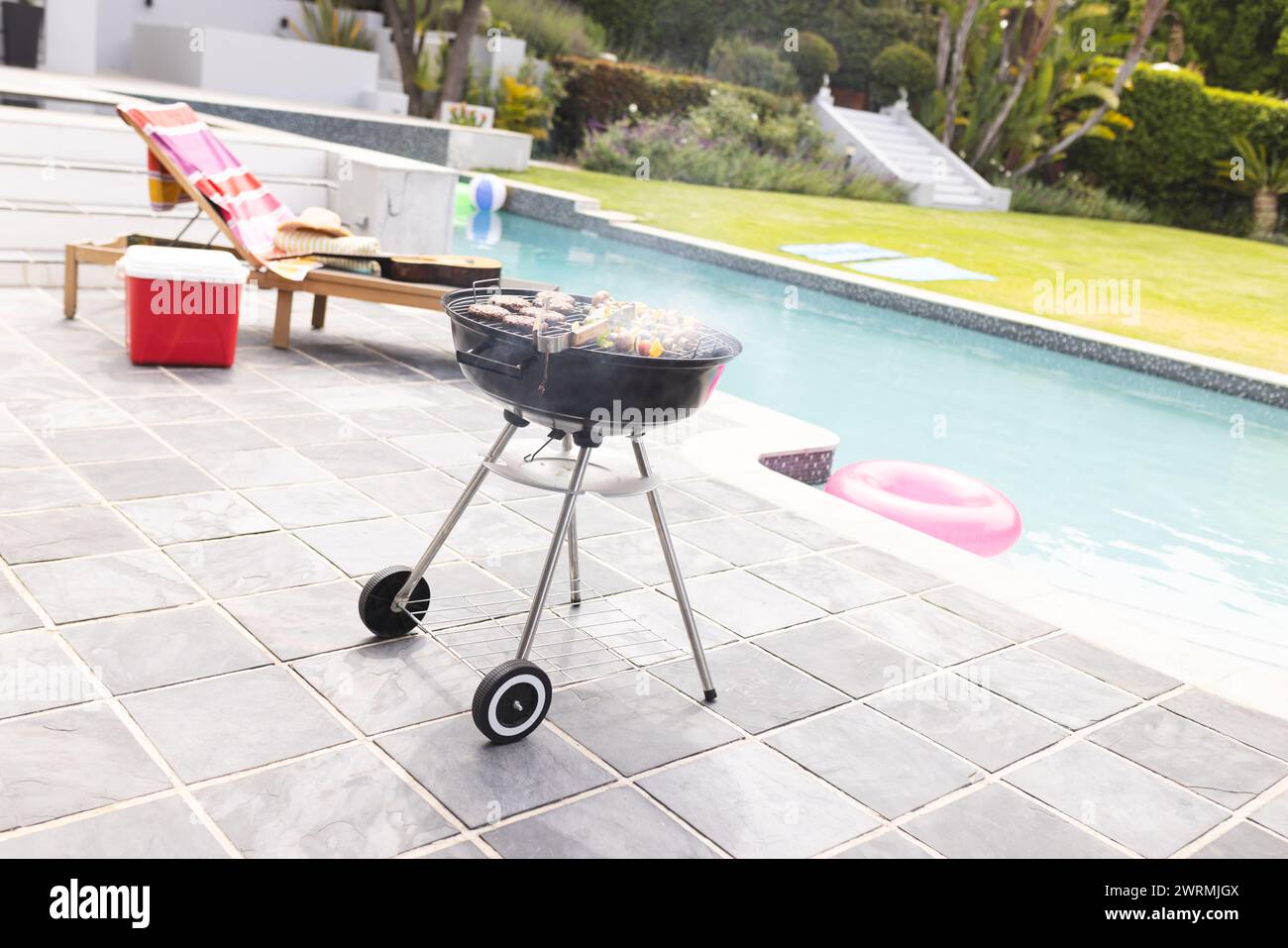 A charcoal grill is smoking beside a pool with a lounge chair and pink ...