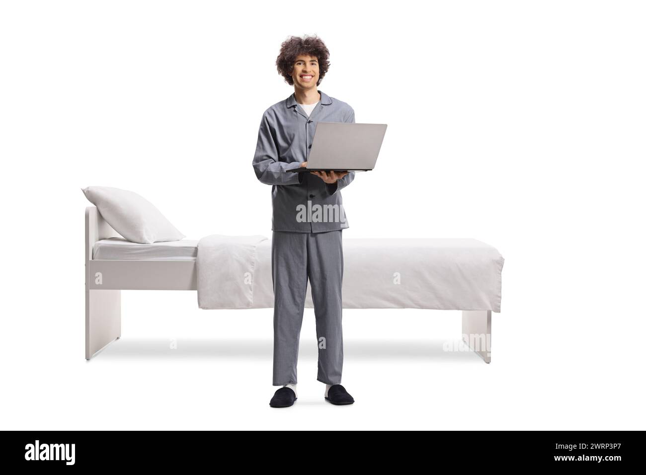 Young man in pajamas standing in front of a bed and working on a laptop ...