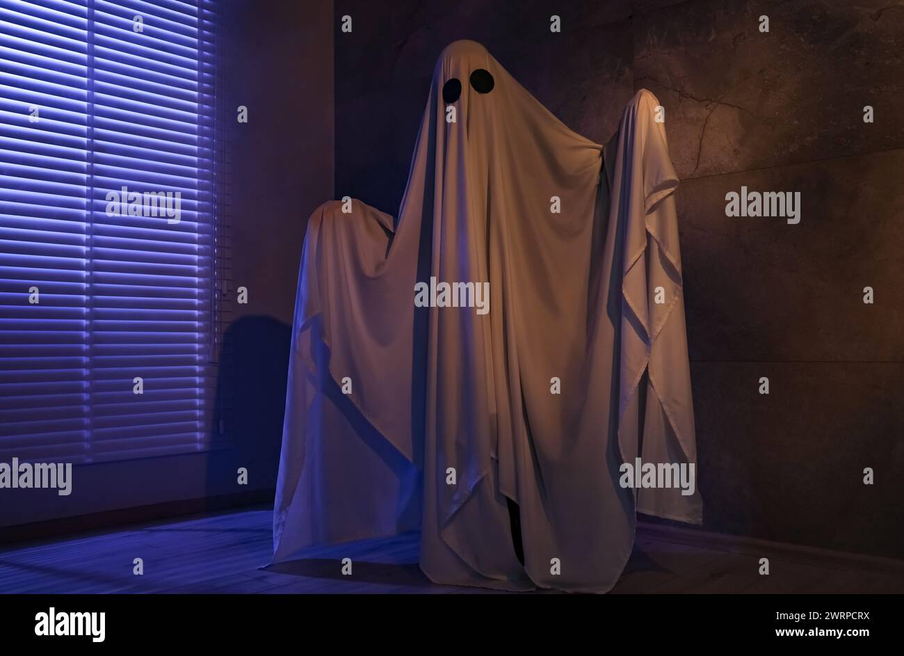 Creepy ghost. Woman covered with sheet near window in color lights ...