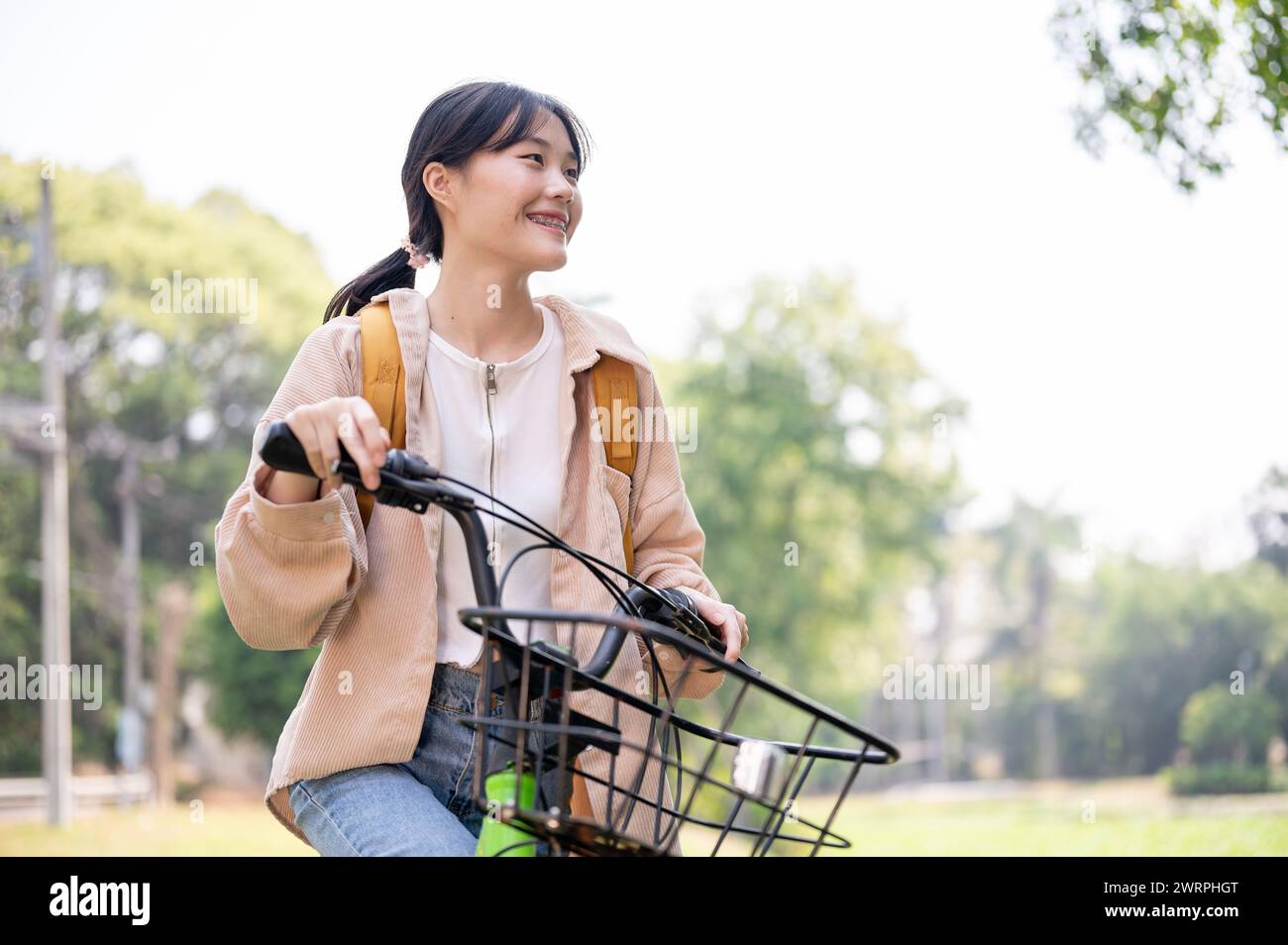 A cheerful young Asian female college student is riding her bike in a green park, on her way to ...