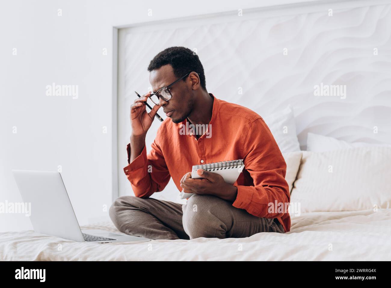A man sits on his bed with a laptop, holding glasses in one hand and a ...