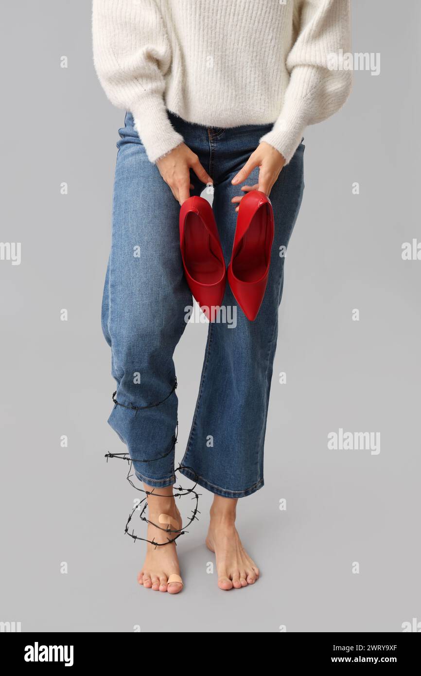 Young woman with plasters and barbed wire on foot holding shoes on grey ...