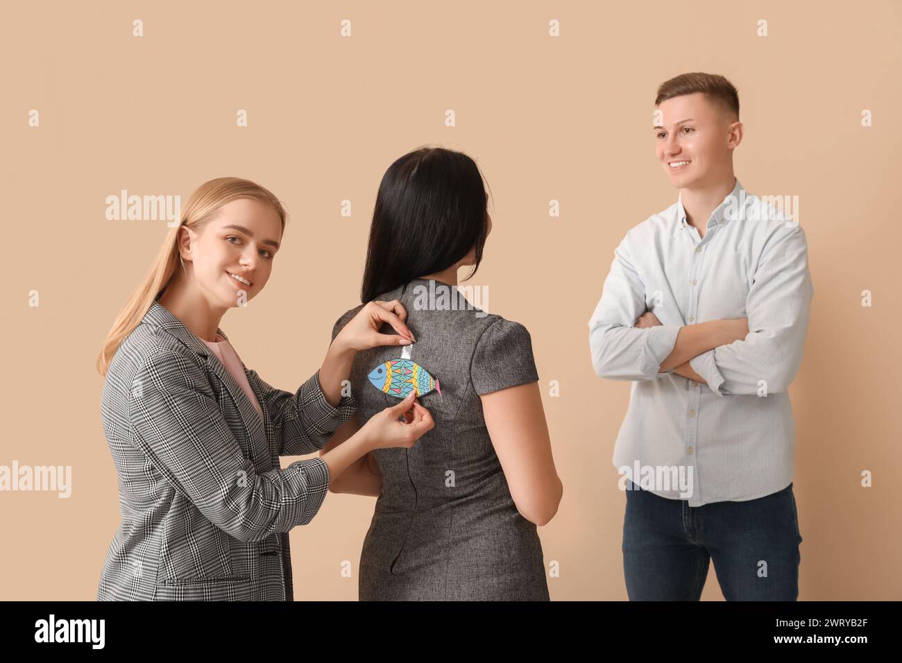Young woman sticking paper fish on her colleague's back against beige ...